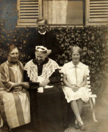 Three ladies seated on a garden bench with reverend vicar standing ...
